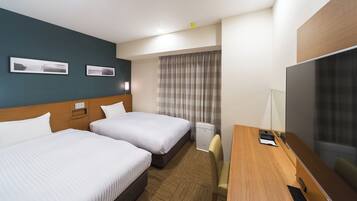 Quarto de hotel com duas camas, uma secretária com televisão de ecrã plano, uma cadeira, uma janela com cortinas e uma luminária fixada na parede.