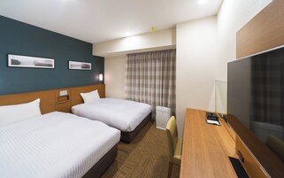 Quarto de hotel com duas camas, uma secretária com televisão de ecrã plano, uma cadeira, uma janela com cortinas e uma luminária fixada na parede.