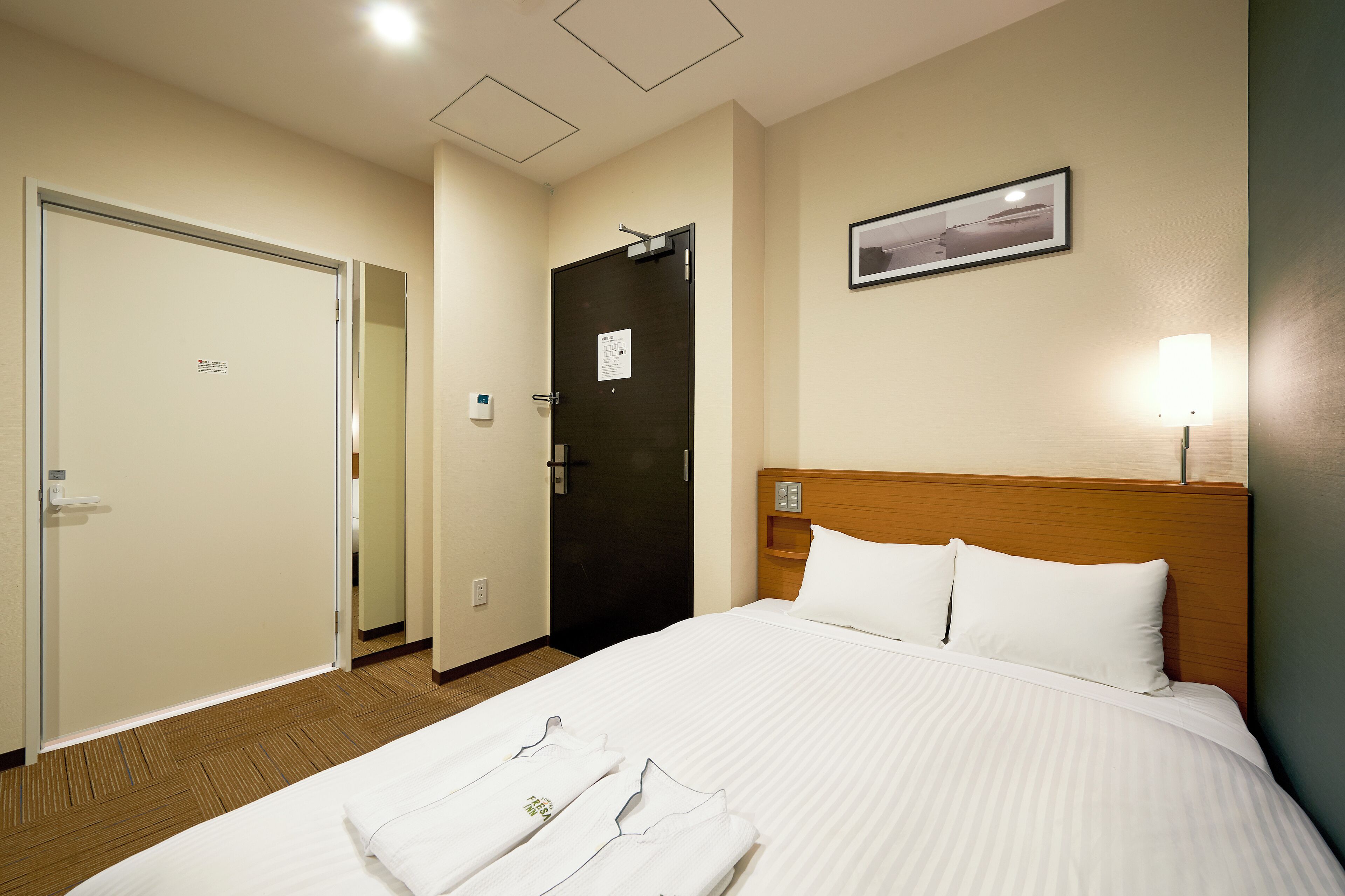 Quarto de hotel com cama, cabeceira de madeira, mesa de cabeceira, luminária, porta e quadro na parede.