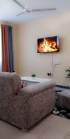 Sala de estar com televisão de tela plana na parede exibindo uma imagem de lareira, ventilador de teto, sofá e uma pequena mesa com uma planta.