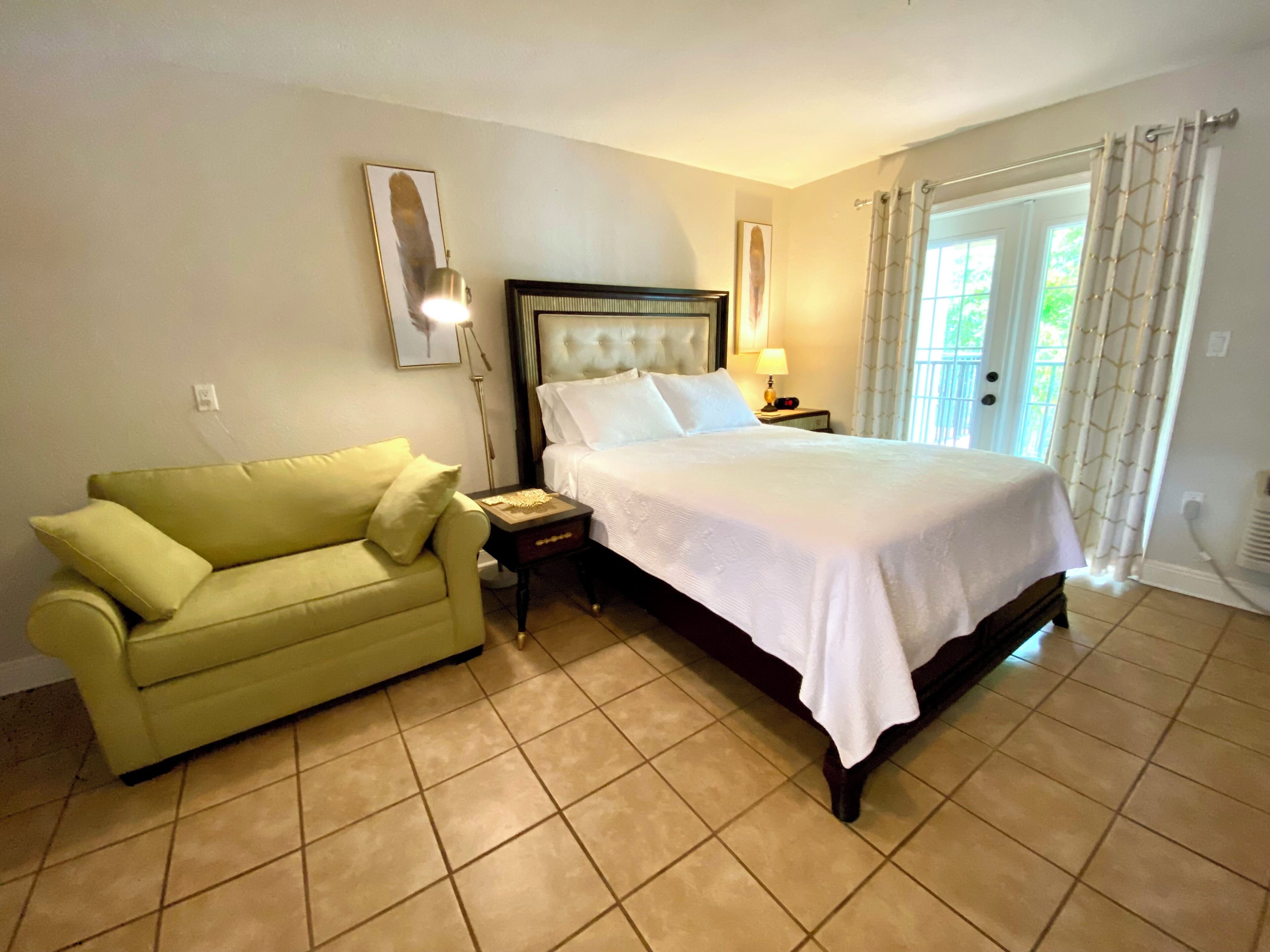 Quarto com cama, sofá, mesa de cabeceira, abajur e janela com cortinas.