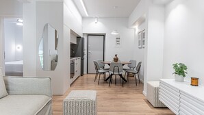 Uma sala de estar moderna e bem iluminada, com sofá, banqueta, mesa de jantar com cadeiras e cozinha ao fundo.