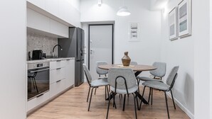 Cozinha moderna com armários brancos, forno embutido, geladeira e área de jantar com mesa redonda e cadeiras estofadas cinzas.