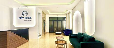 Um saguão de hotel moderno com um balcão de recepção, uma escadaria, um sofá verde e uma placa com a inscrição 'MAY HOUSE mini hotel'.