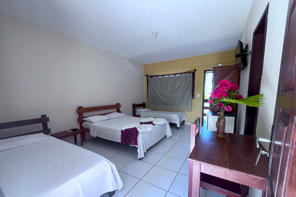 Quarto de hotel com duas camas de solteiro, uma escrivaninha de madeira, um espelho e um buquê de flores.