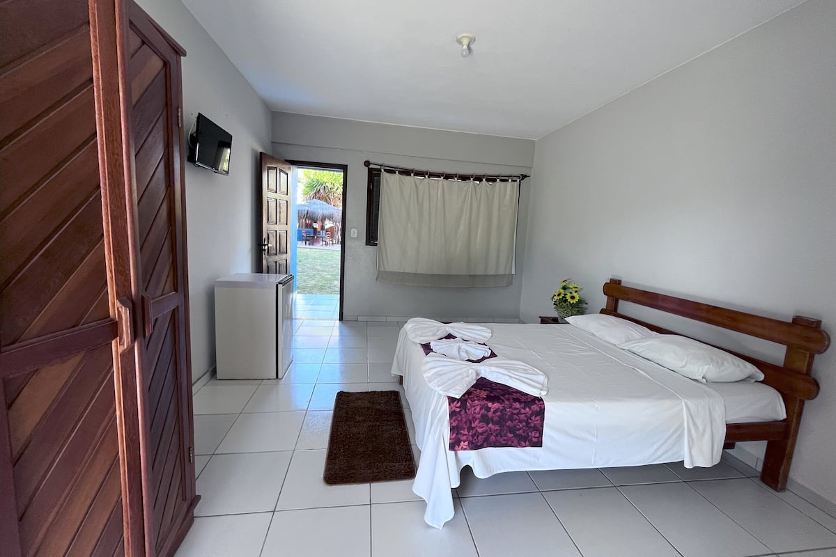Quarto com cama, guarda-roupa de madeira, uma secretária pequena e televisão na parede.