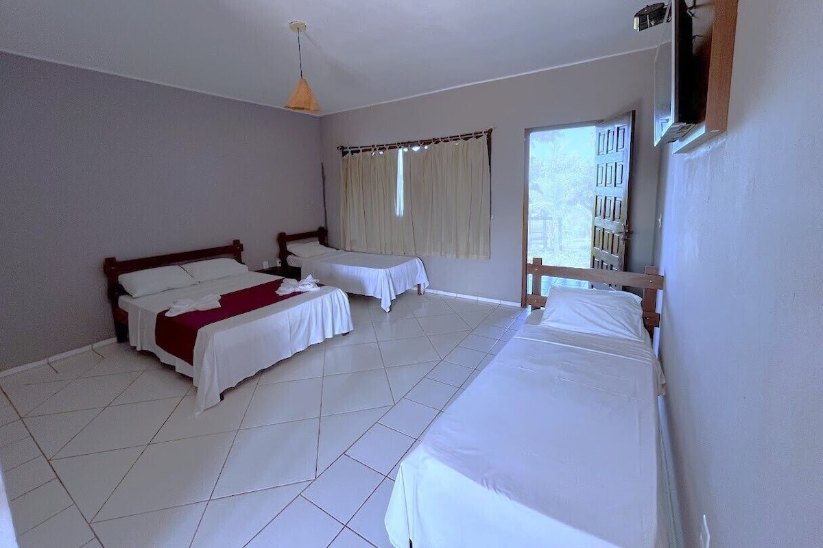 Quarto de hotel com três camas de solteiro, televisão fixada na parede e janela com cortinas.
