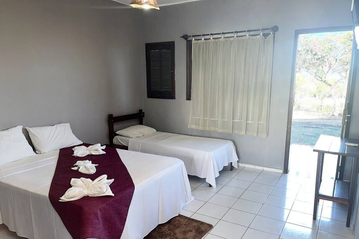 Quarto de hotel com duas camas, uma janela com cortinas leves e um ventilador de teto, com vista para o exterior.