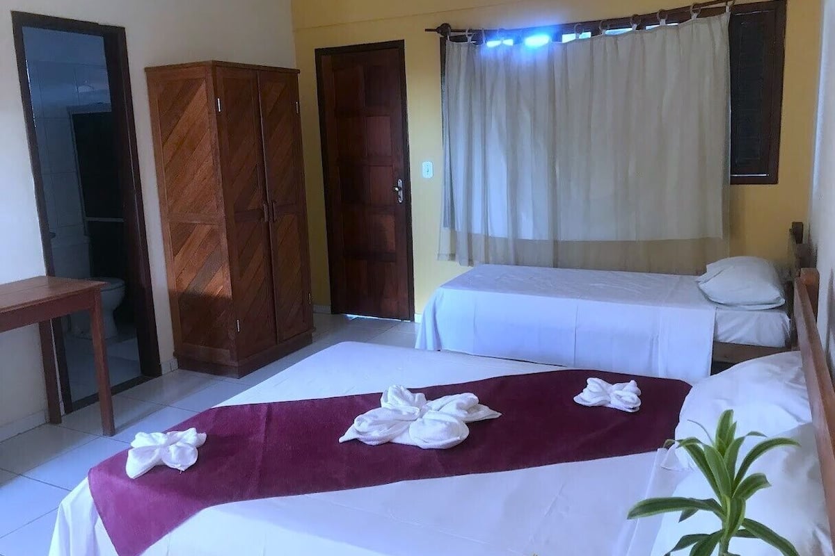 Quarto de hotel com duas camas, um guarda-roupa, banheiro e uma pequena mesa.