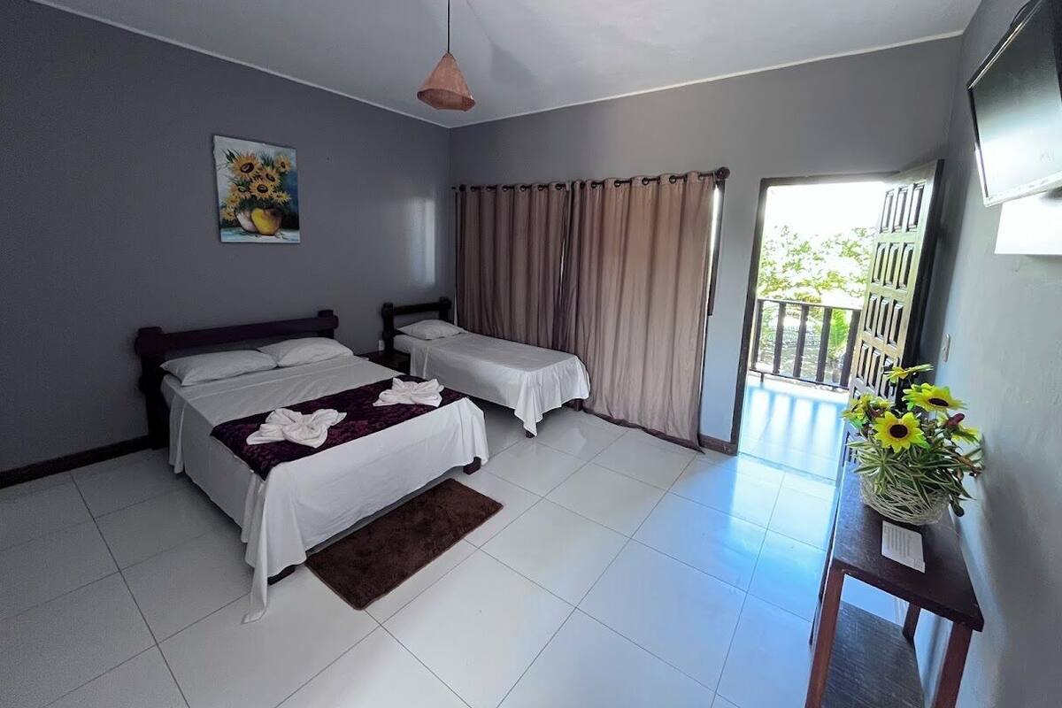 Quarto de hotel com duas camas, uma mesinha com um vaso de flores, um quadro na parede e uma varanda com cortinas.