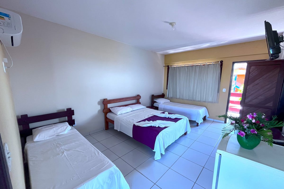 Quarto de hotel com três camas de solteiro, uma escrivaninha, televisão e janela com cortinas.