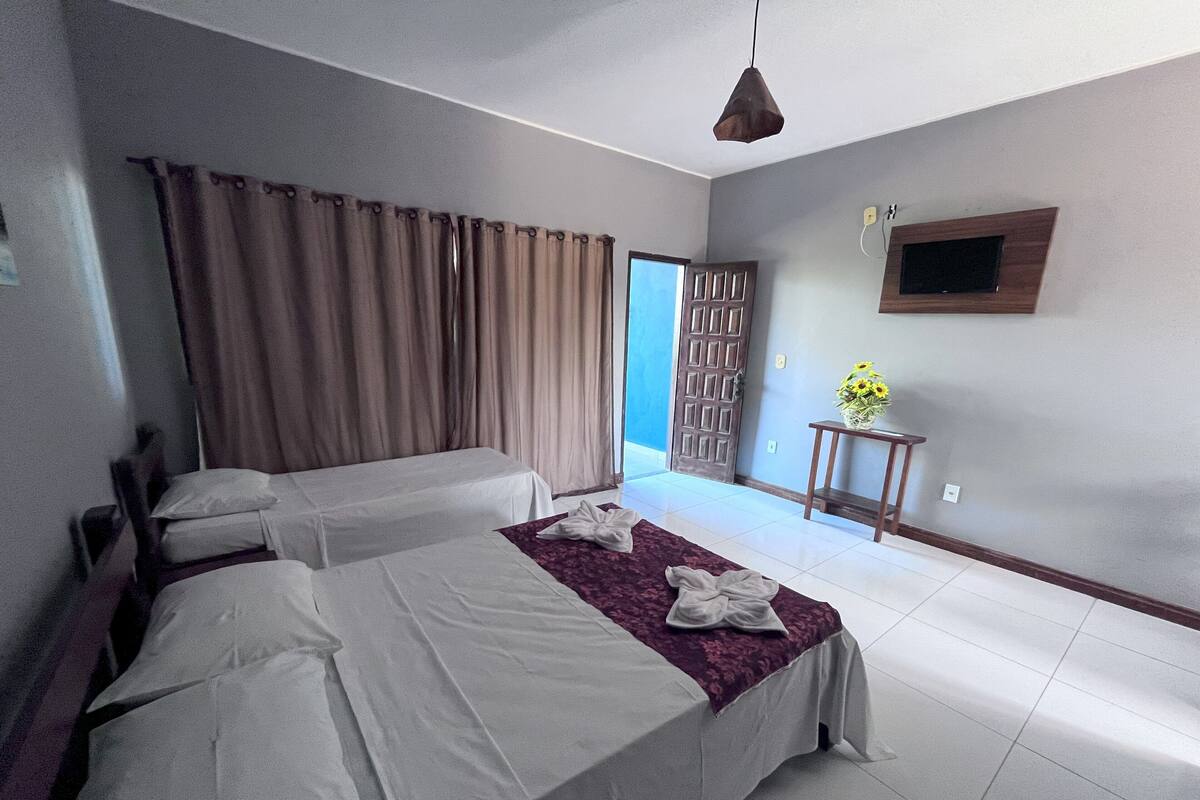 Quarto de hotel com duas camas, televisão presa na parede, uma mesinha com um vaso de flores e uma porta que dá para uma varanda.
