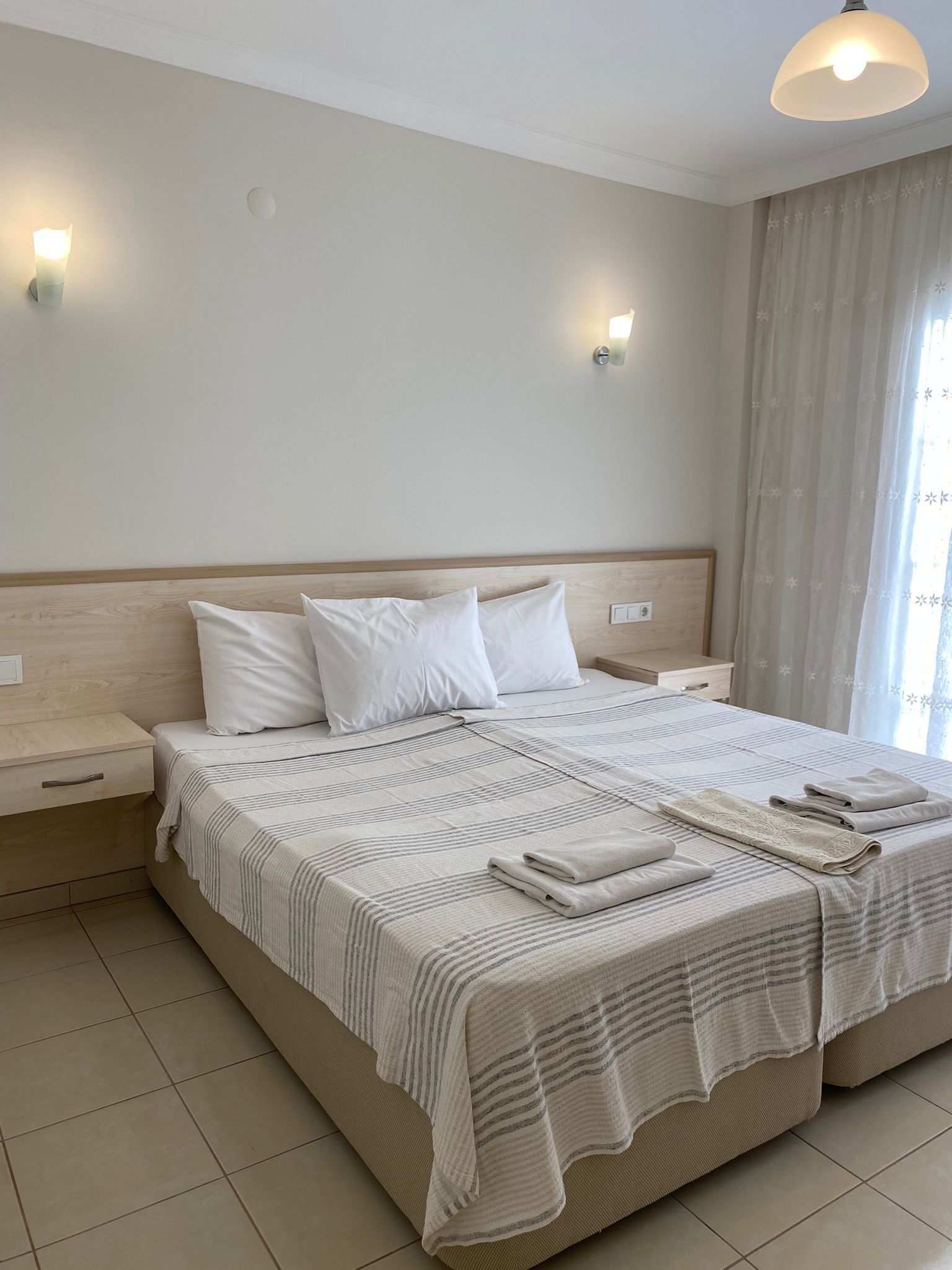 Uma cama bem arrumada com lençóis brancos e manta listrada, duas mesas de cabeceira com luminárias, e uma janela grande com cortinas.