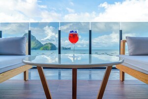 Uma mesa de vidro com uma bebida vermelha, piso de madeira e vista para o mar e montanhas através de grandes janelas.