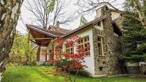 Uma casa com fachada de pedra, varanda de madeira e um jardim bem cuidado com folhagem avermelhada.