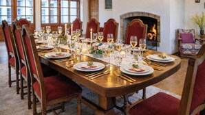 Uma sala de jantar com uma mesa comprida posta para refeição, cadeiras estofadas vermelhas, uma lareira e vista para o exterior através de grandes janelas.