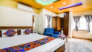 Quarto de hotel com uma cama grande, uma poltrona azul, uma mesa pequena com uma garrafa e uma janela com cortinas.