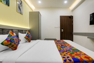 Quarto de hotel com cama, almofadas coloridas, mesa de cabeceira, guarda-roupa e porta.