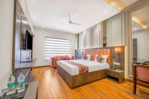 Quarto de hotel moderno com uma cama grande, televisão de tela plana, piso de madeira, ventilador de teto e janela com persianas.