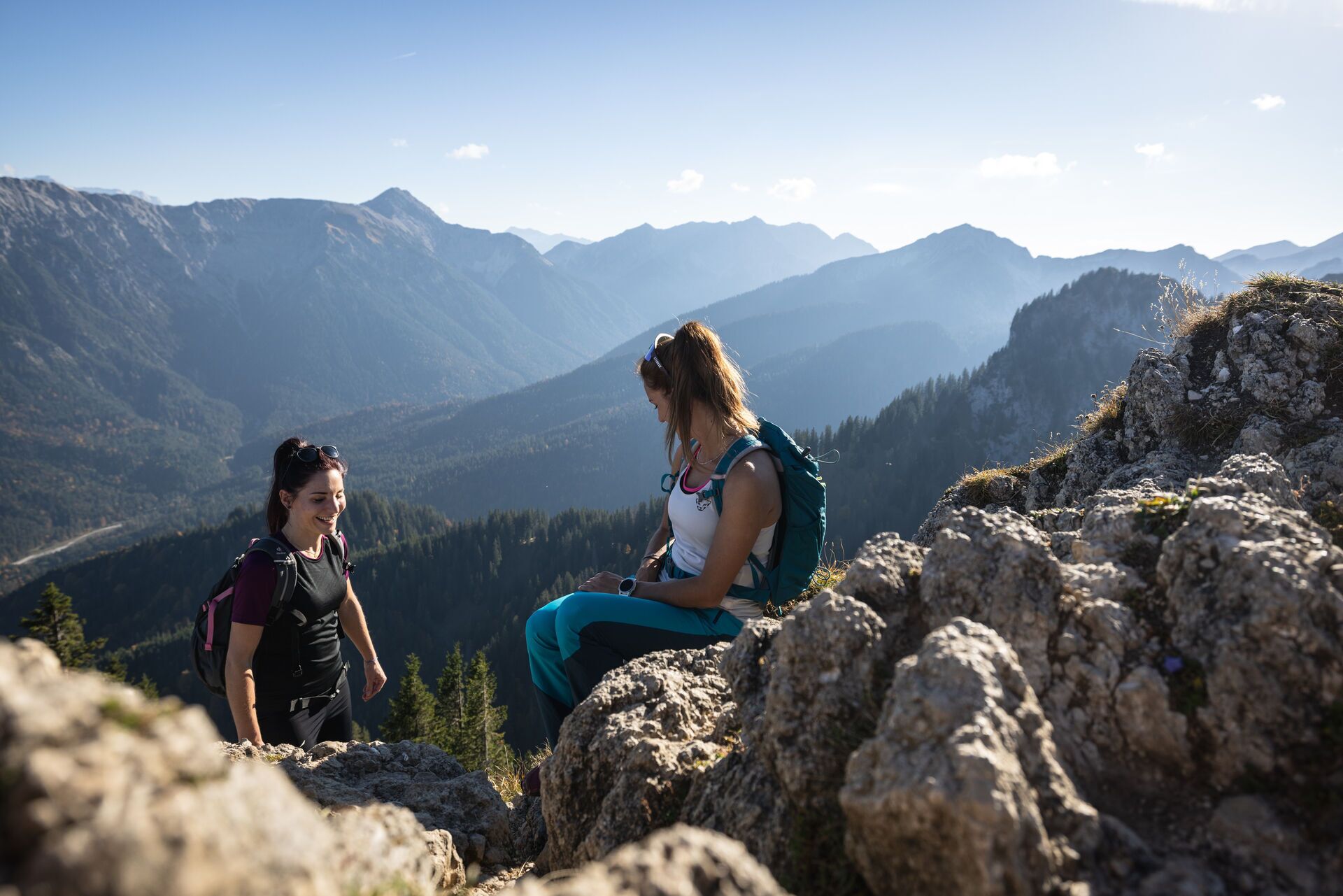 Duas pessoas sentadas em um afloramento rochoso numa montanha, com vista para montanhas e vales distantes.
