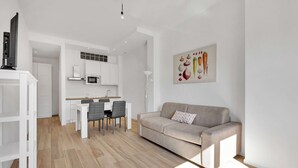 Uma sala de estar moderna com sofá, área de refeições com cadeiras, cozinha com armários brancos e uma TV de tela plana.