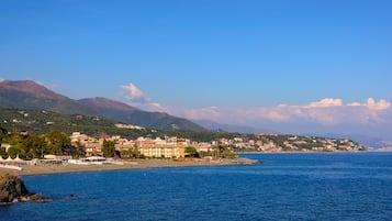 Uma vila costeira com edifícios ao longo da praia, um afloramento rochoso na água e montanhas ao fundo.
