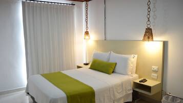 Cama bem arrumada com roupa de cama branca e manta verde, mesa de cabeceira com abajur e lixeira.