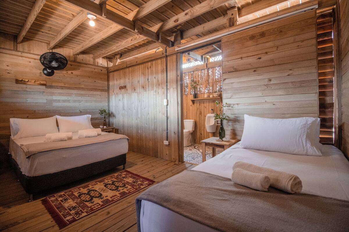 Quarto estilo cabana de madeira com duas camas, teto de madeira e uma pequena mesa com abajur.