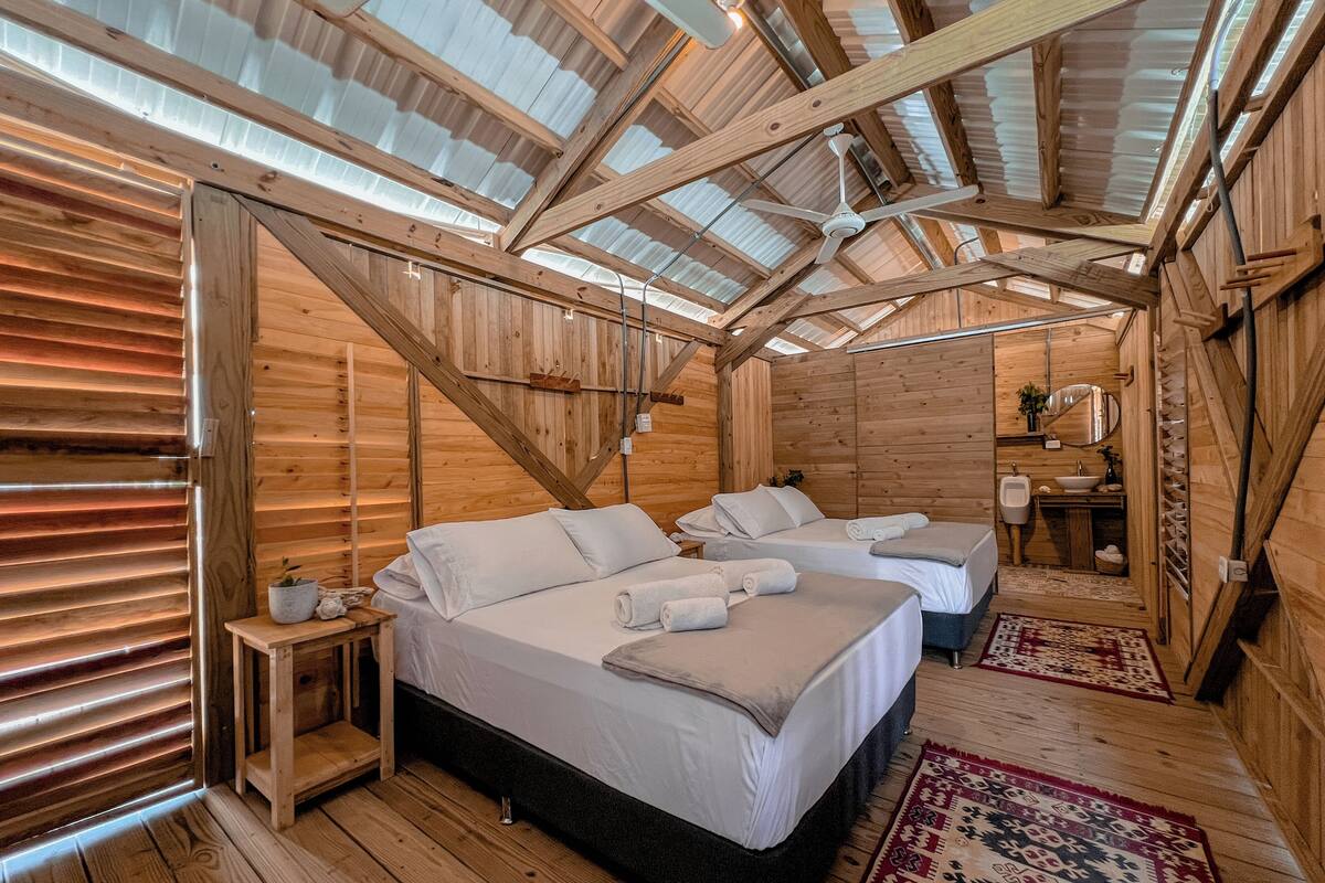 Uma cabana de madeira com teto inclinado, uma cama com almofadas e toalhas, uma mesa pequena, um espelho e uma pia.