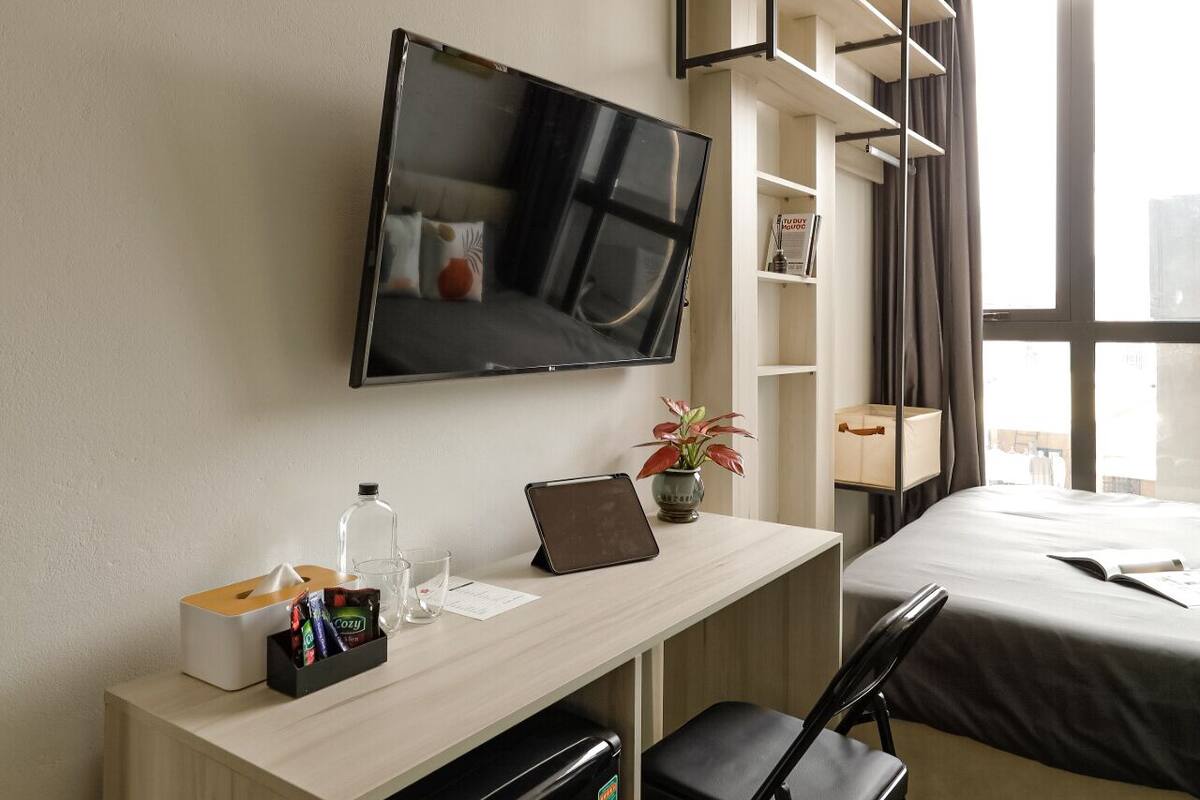 Quarto de hotel moderno com televisão de tela plana na parede, uma escrivaninha com cadeira, uma cama e uma janela com cortinas.