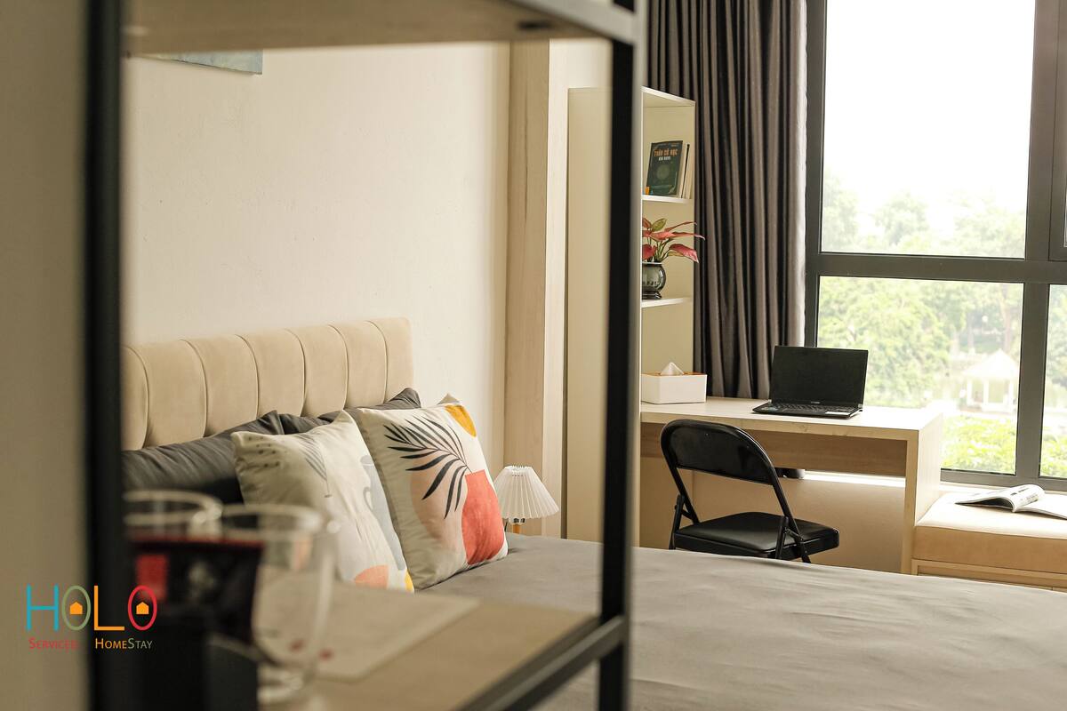 Quarto de hotel com cama, escrivaninha com um laptop, cadeira e janela com vista para o verde.