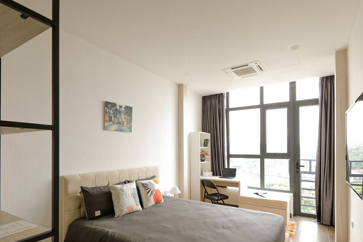 Quarto de hotel moderno com uma cama grande, uma escrivaninha com computador e vista para o exterior através de amplas janelas.