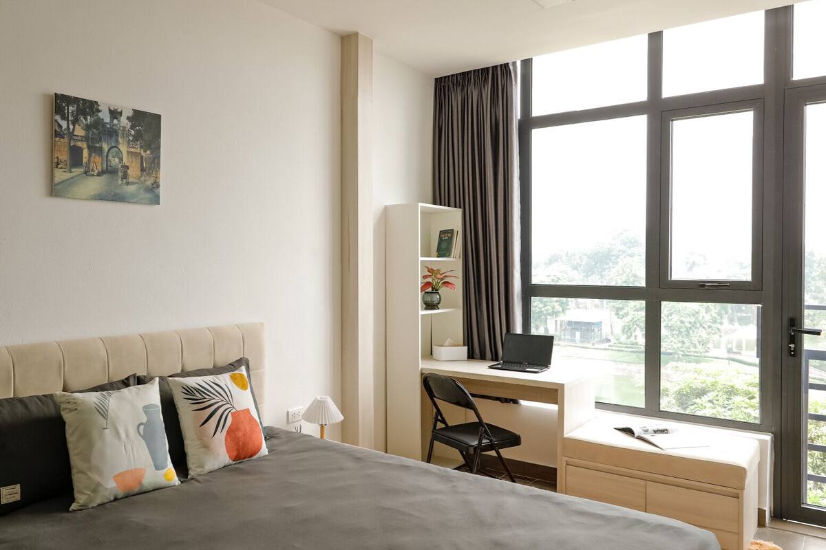 Quarto moderno com uma cama grande, uma escrivaninha e uma cadeira. Há uma janela com cortinas, um quadro na parede e um laptop na escrivaninha.