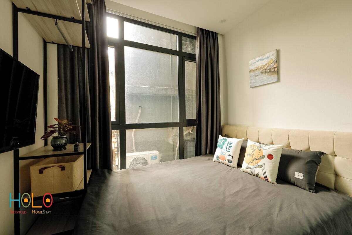 Quarto de hotel com uma cama grande, televisão em suporte, escrivaninha com planta e janela com cortinas.