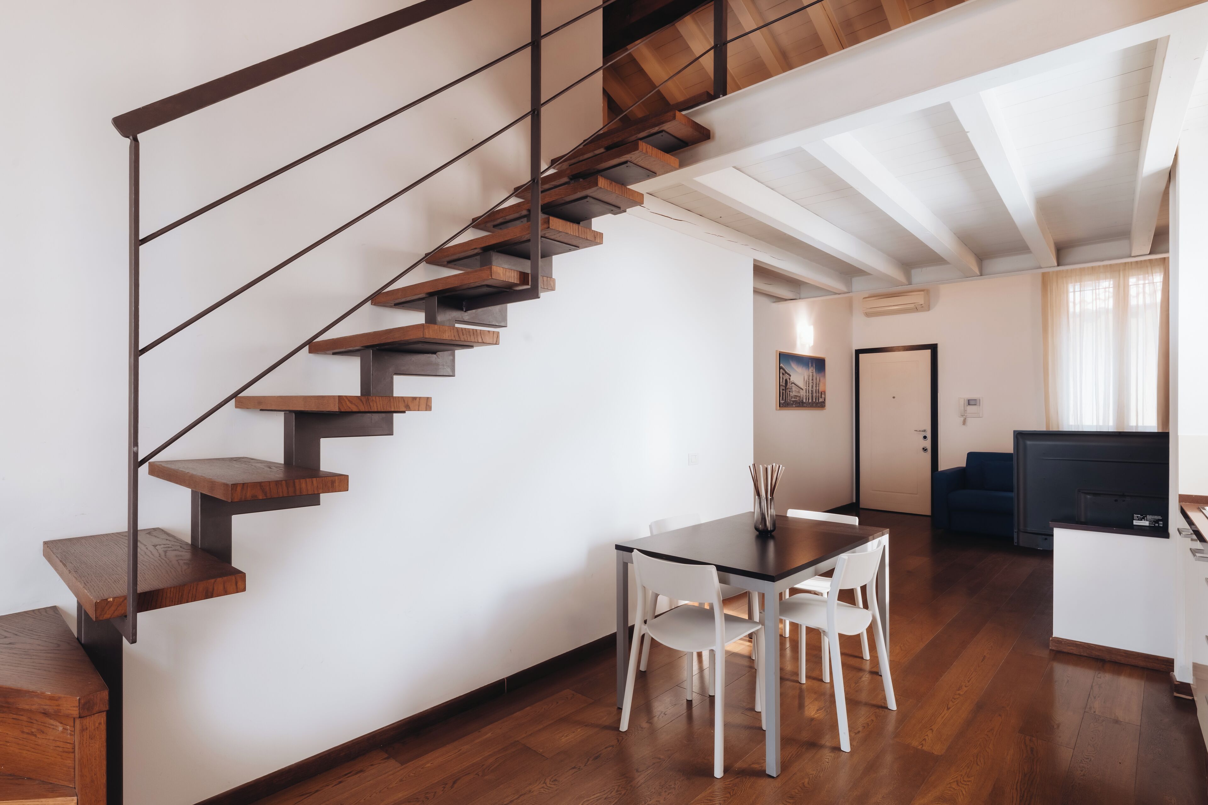 Um interior moderno com escada de madeira, sala de jantar com cadeiras brancas e área de estar com sofá e televisão.