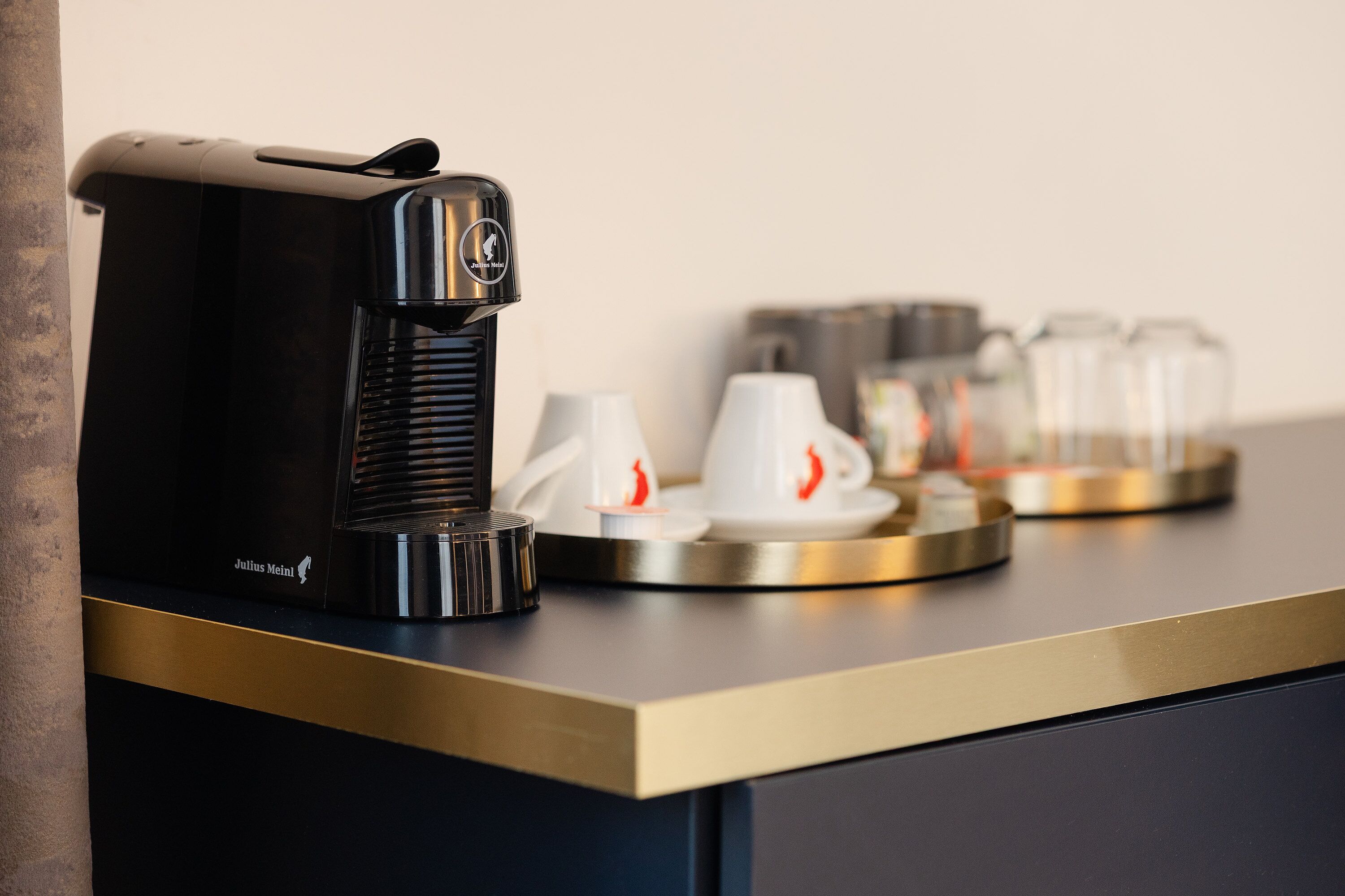 Uma cafeteira moderna sobre um balcão com detalhe dourado, uma chávena branca e um jarro de vidro.