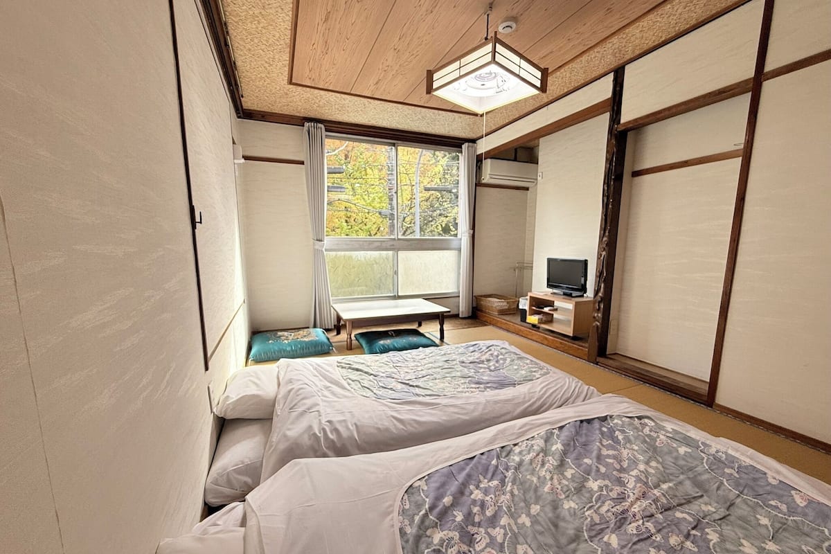 Quarto com cama, escrivaninha, televisão e janela com vista para árvores.