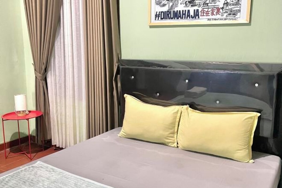 Quarto de hotel com cama, cortinas, uma pequena mesa com abajur e um quadro na parede.