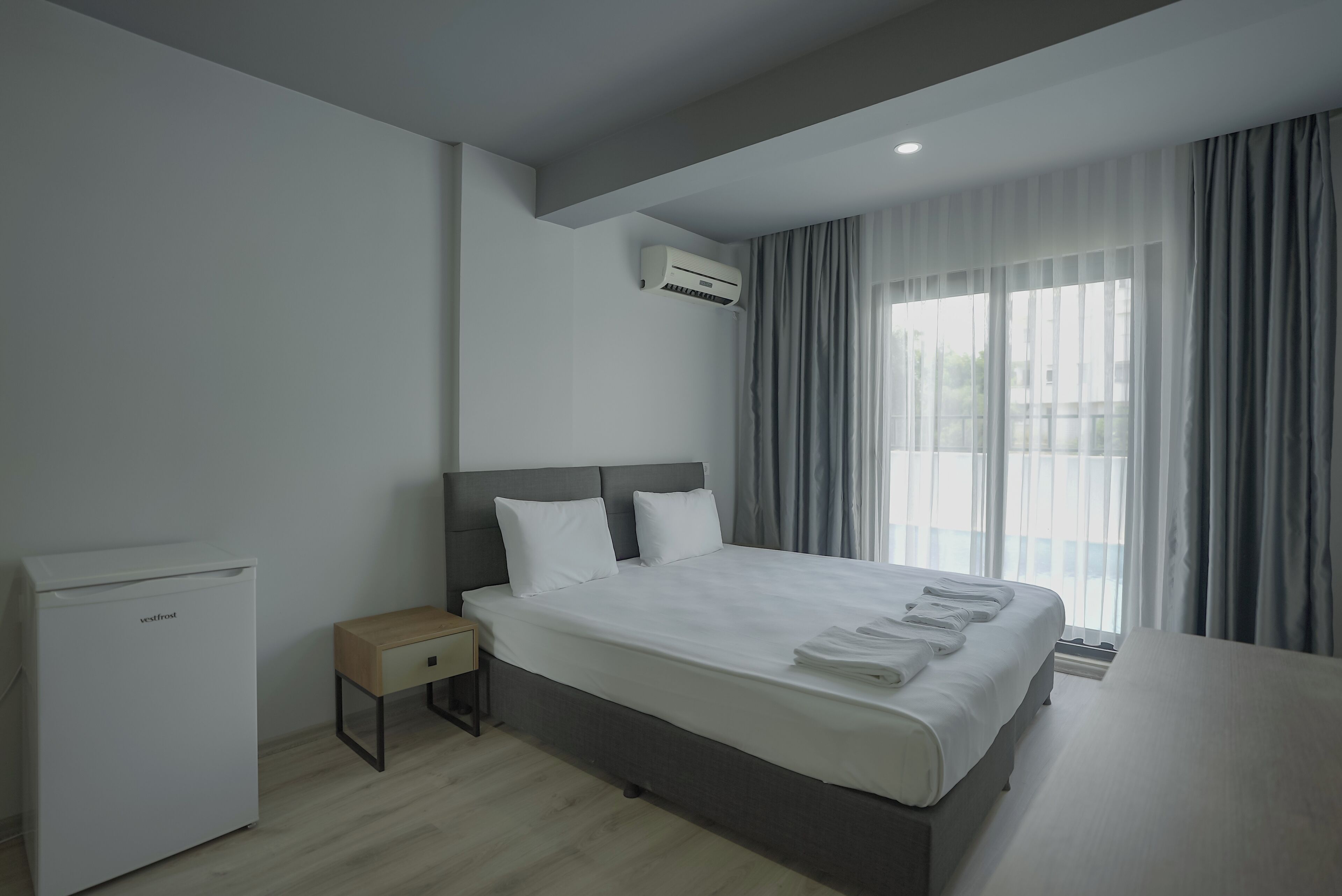 Quarto de hotel moderno com uma cama grande, mesa de cabeceira, um pequeno frigorífico e uma janela com cortinas.