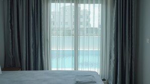 Quarto de hotel com cama, vista para uma piscina através de cortinas leves e varanda com guarda-corpo.