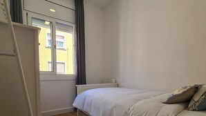 Quarto com cama, mesa de cabeceira, janela com cortinas e vista para um edifício amarelo.