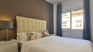 Quarto com uma cama grande, mesa de cabeceira com abajur e vista para edifícios pela janela.