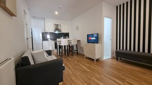Uma sala de estar moderna com um sofá preto, uma televisão e uma cozinha ao fundo.
