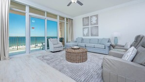 Uma sala de estar espaçosa com uma grande porta de vidro deslizante que dá para uma varanda com vista para a praia. Há um sofá cinzento, uma mesa de centro de vime e uma poltrona cinza.