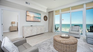 Uma sala de estar ampla com uma grande janela com vista para uma praia, um sofá confortável, uma mesa de centro de vime e um armário branco com painéis decorativos.
