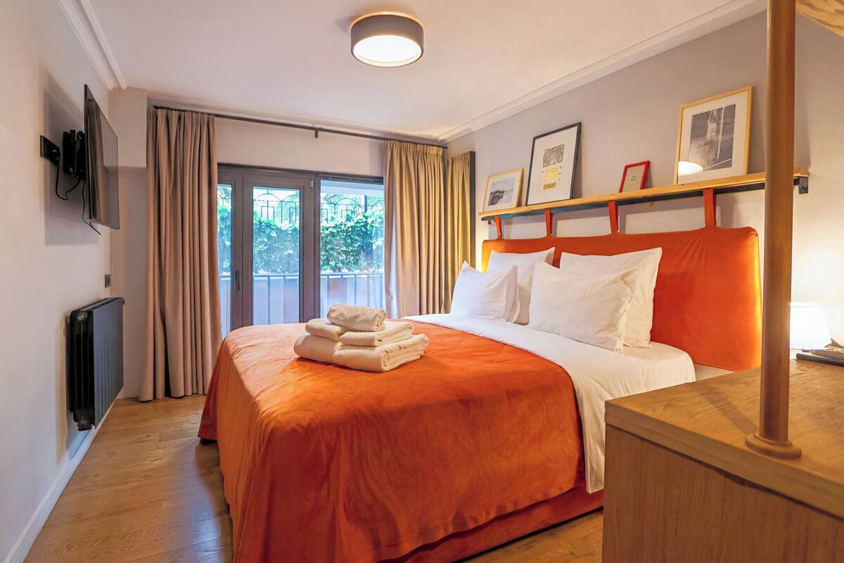 Quarto de hotel com cama, televisão, janela com cortinas e vista para vegetação lá fora.