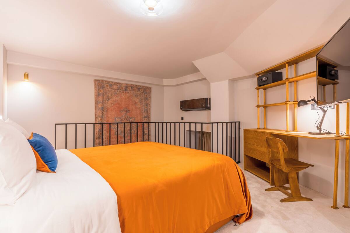 Quarto com cama, escrivaninha, cadeira, televisão e prateleira com caixas organizadoras.
