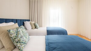 Quarto de hotel com duas camas, cabeceira azul e uma janela grande com cortinas finas.