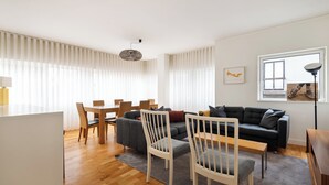 Uma sala de estar moderna com um sofá, cadeiras, uma mesa de jantar e uma janela grande com persianas.