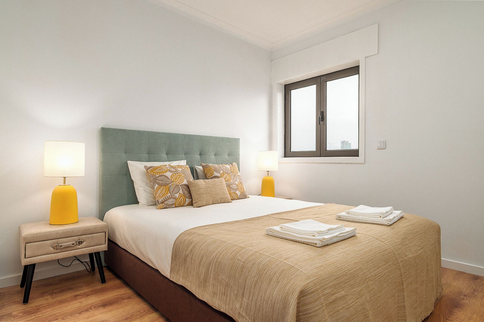 Quarto com uma cama, duas mesas de cabeceira, luminárias, uma janela e vista para a cidade.
